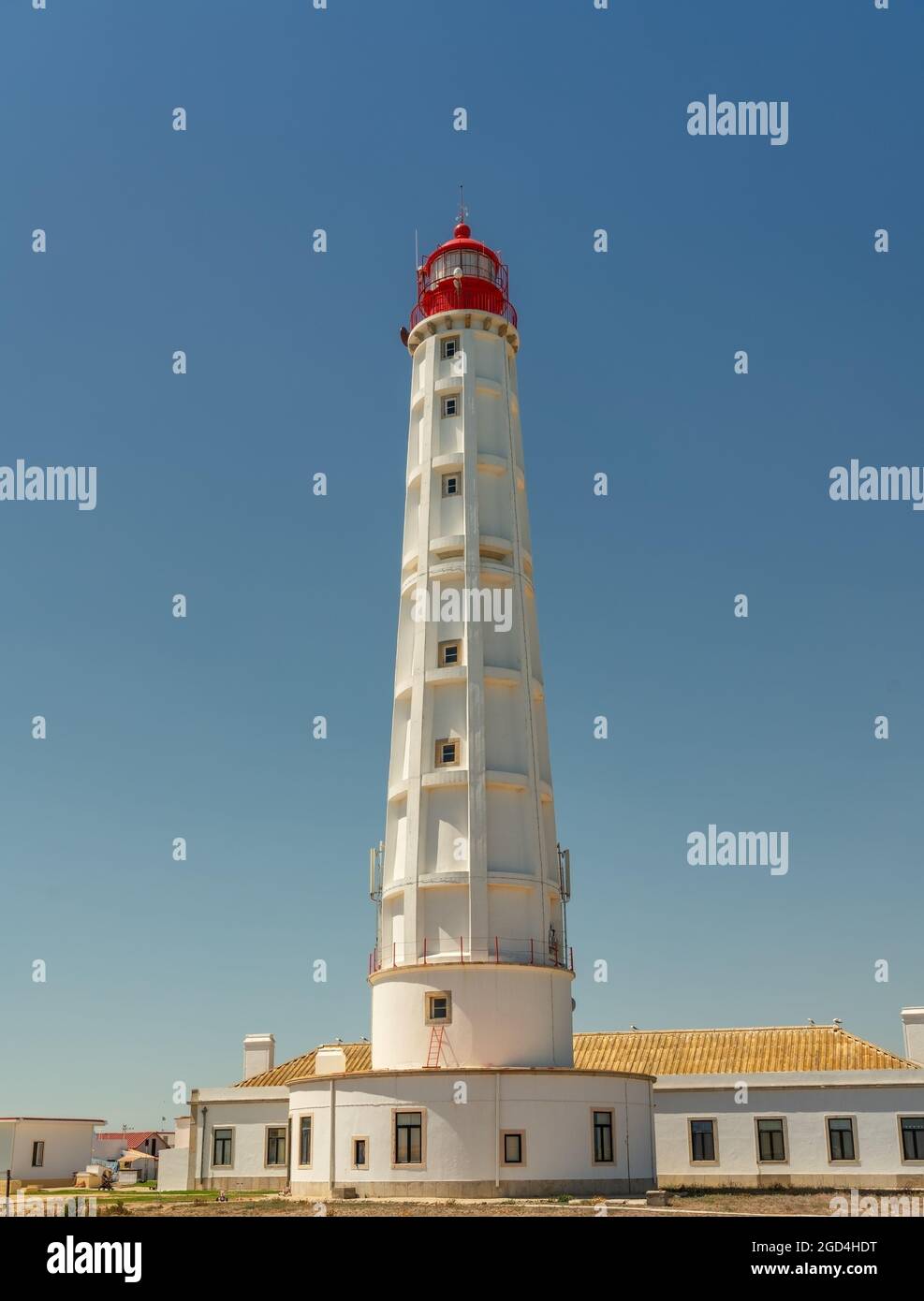 A white lighthouse with a red top, against the blue sky. On a sandy ...