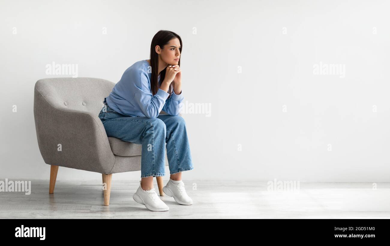 Portrait of annoyed young lady sitting in armchair against white studio ...