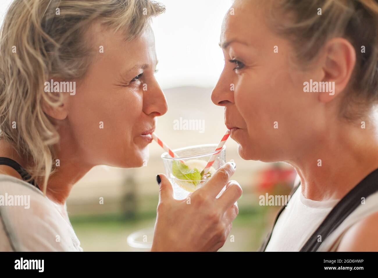 Two cheerful girlfriends drink beverage from one glass using straws ...