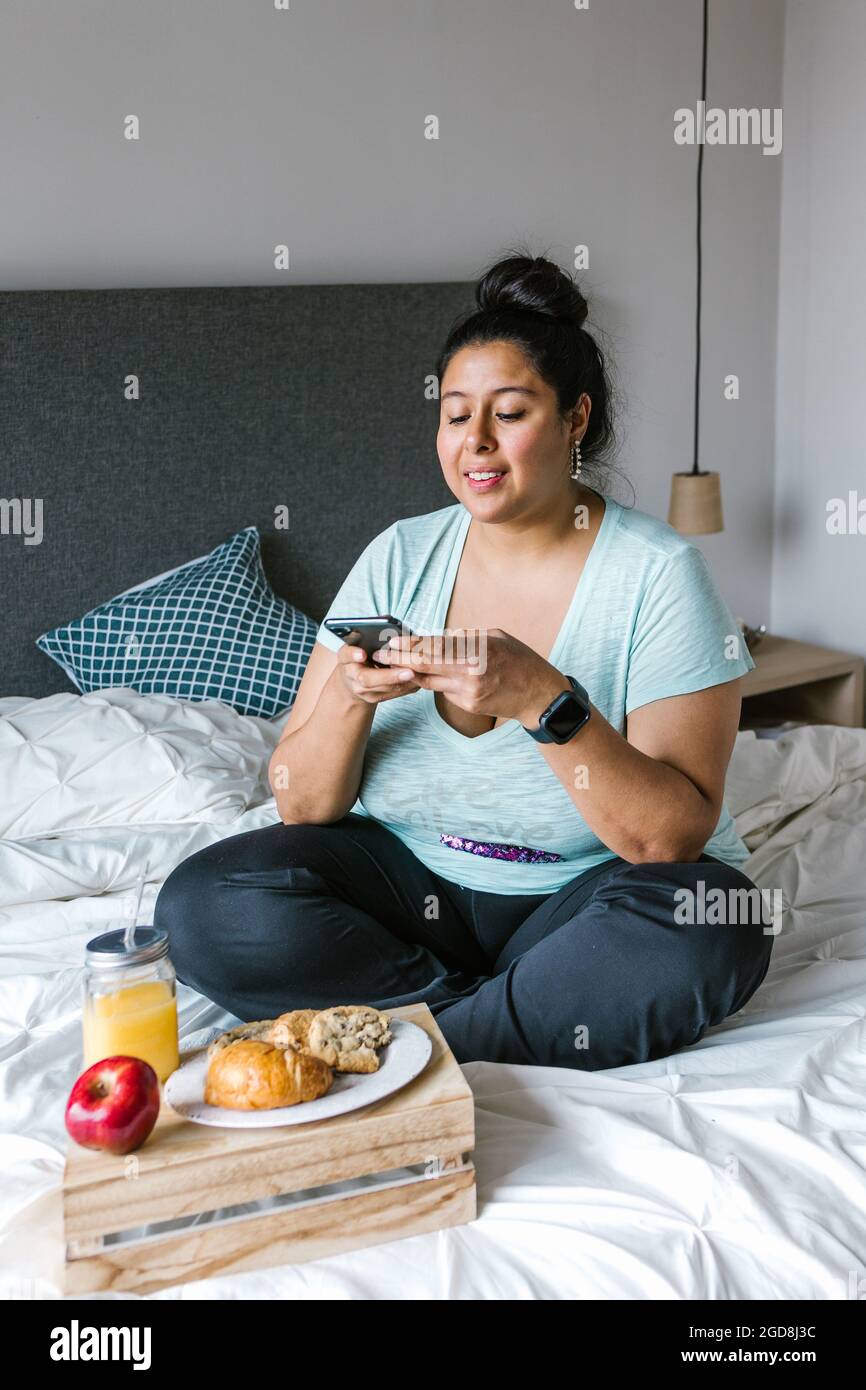 Young mexican plus size woman sitting in bed using phone while having ...