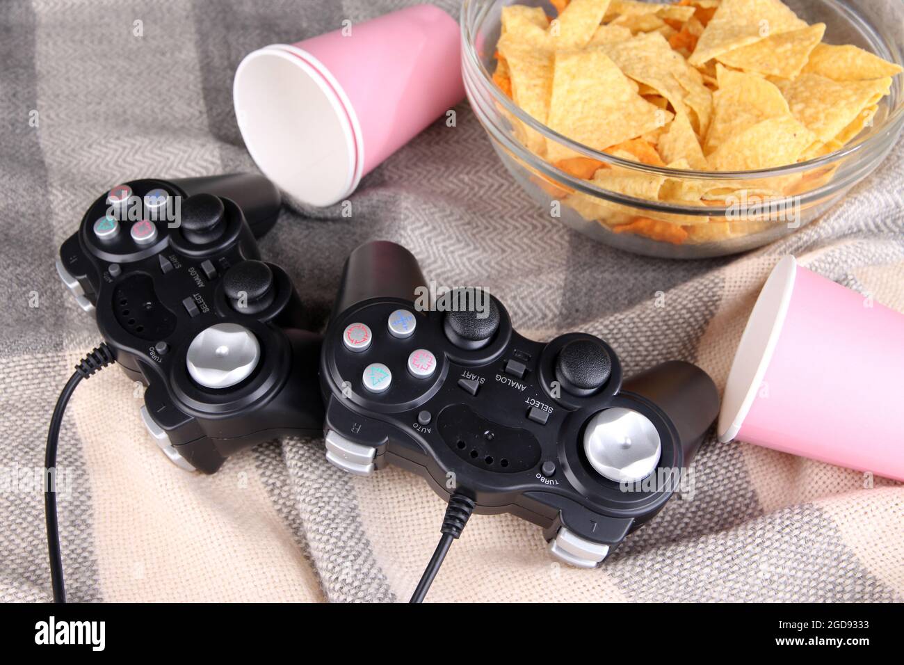 Black game controllers and bowl with snacks on color plaid background ...