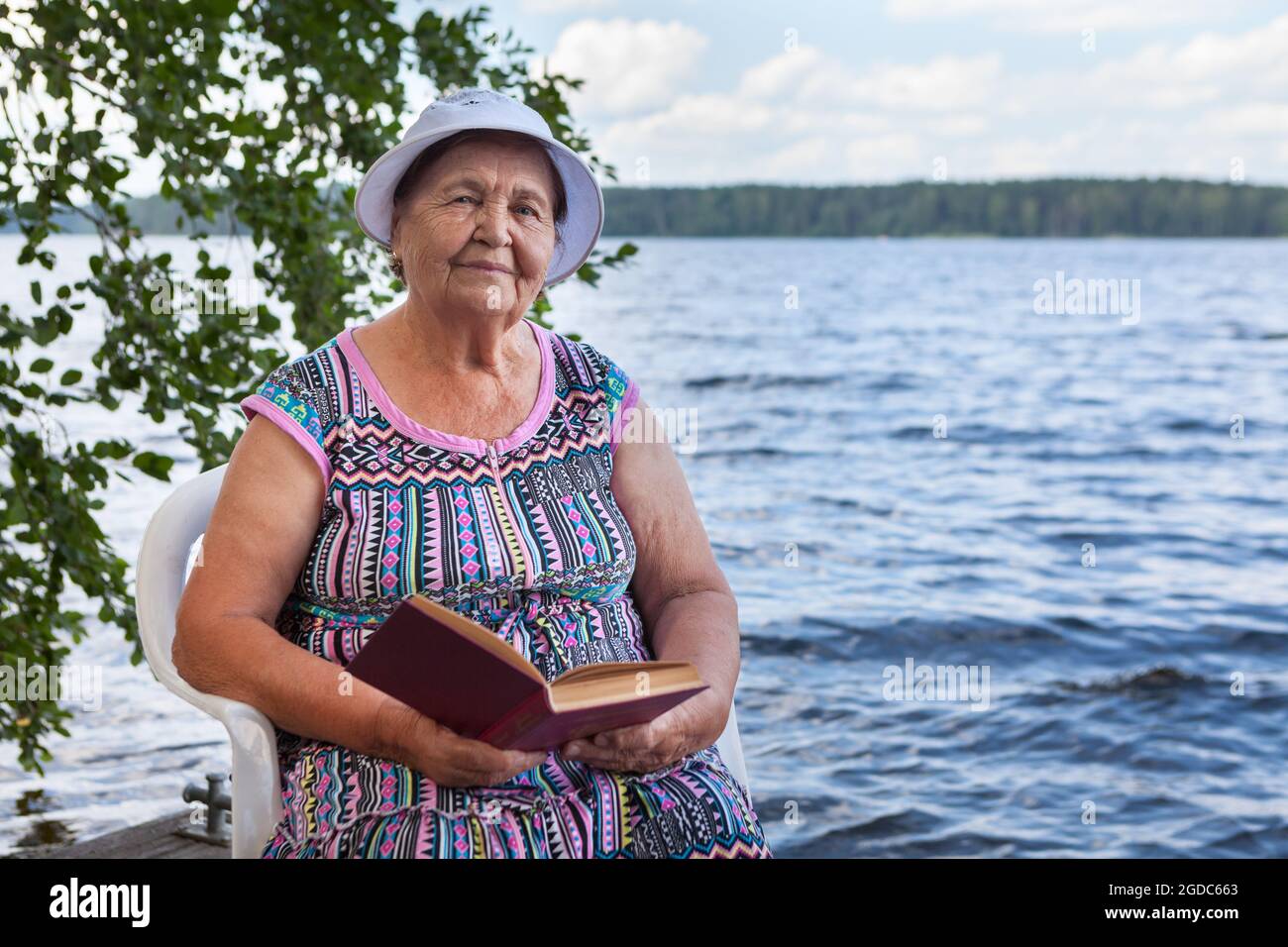 Old lady reading book sitting at the plastic chair in forest lake shore ...