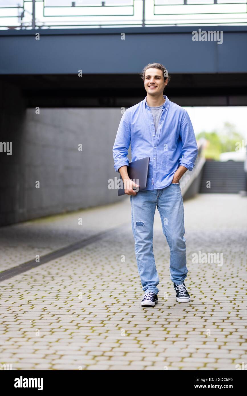 Portrait of a young serious man holding laptop computer while standing ...