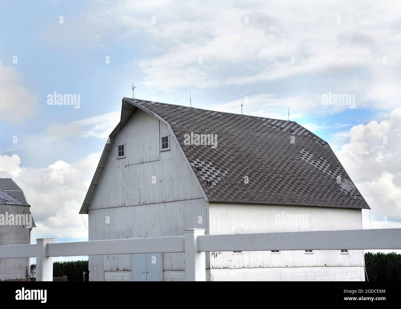 White wooden barn with shingle roof sits behind a white fence. Sunny ...