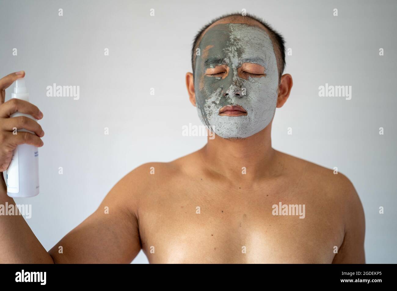 Thai male with a clay facial mask spraying his face isolated on a gray ...