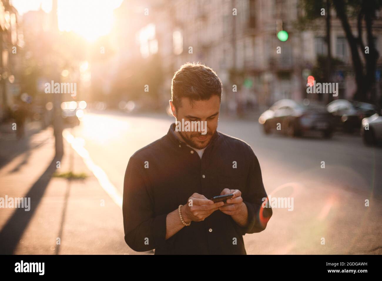 Happy man using smart phone standing on street in city during summer ...