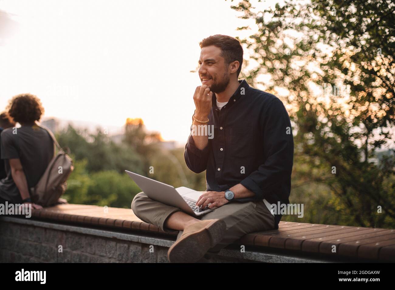 Happy thoughtful man using laptop computer sitting on bench in park in ...