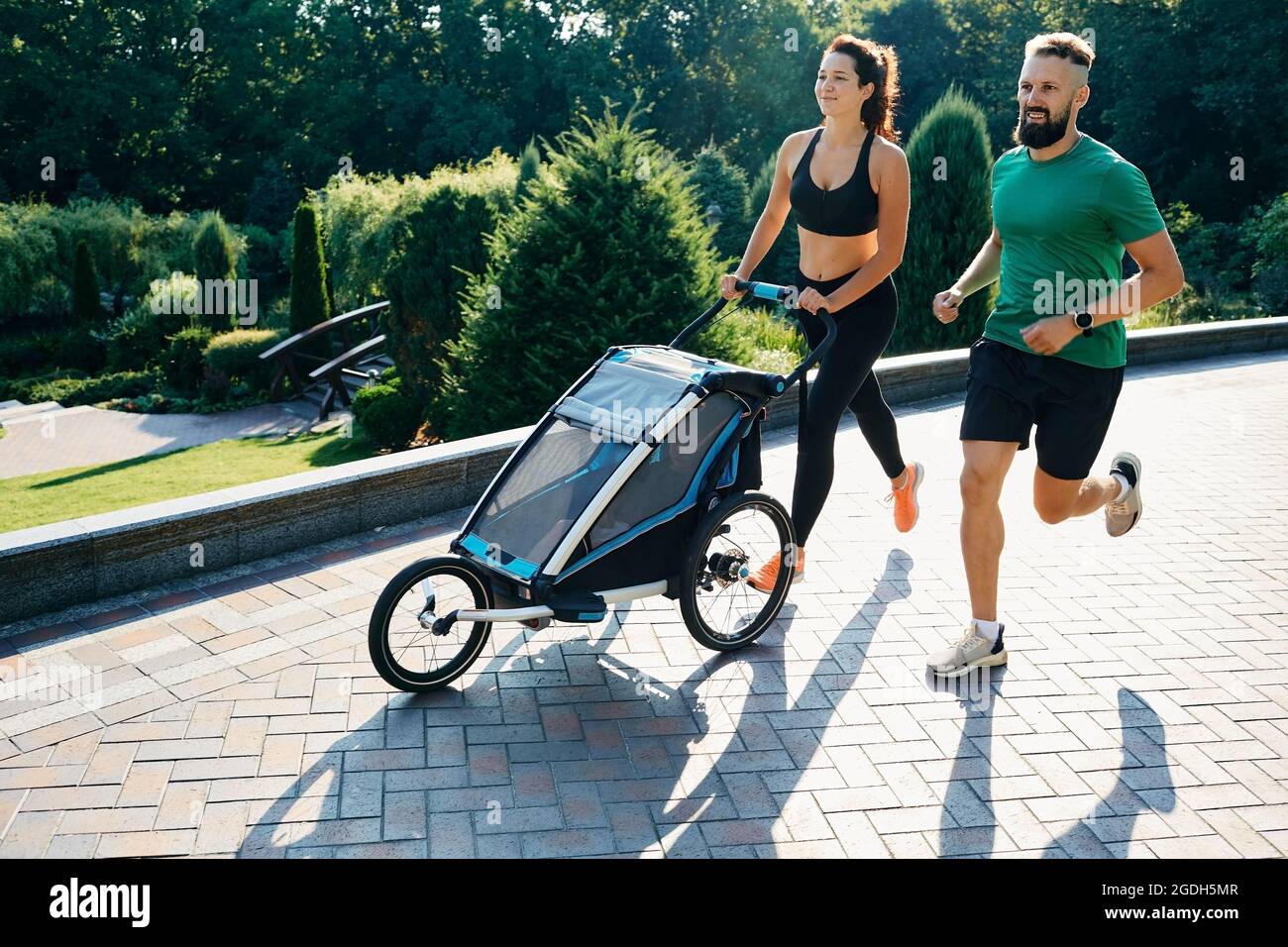 running couple with child in jogging stroller in public park at sunrise ...