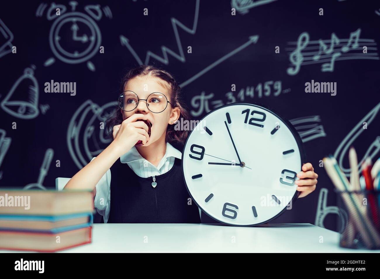Pupil girl with big clock eating apple at the black chalkboard in ...