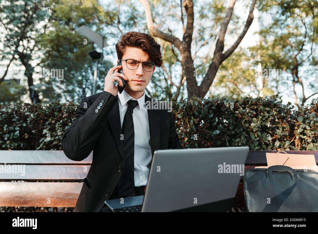 Serious professional young Caucasian guy sitting on a bench using his ...