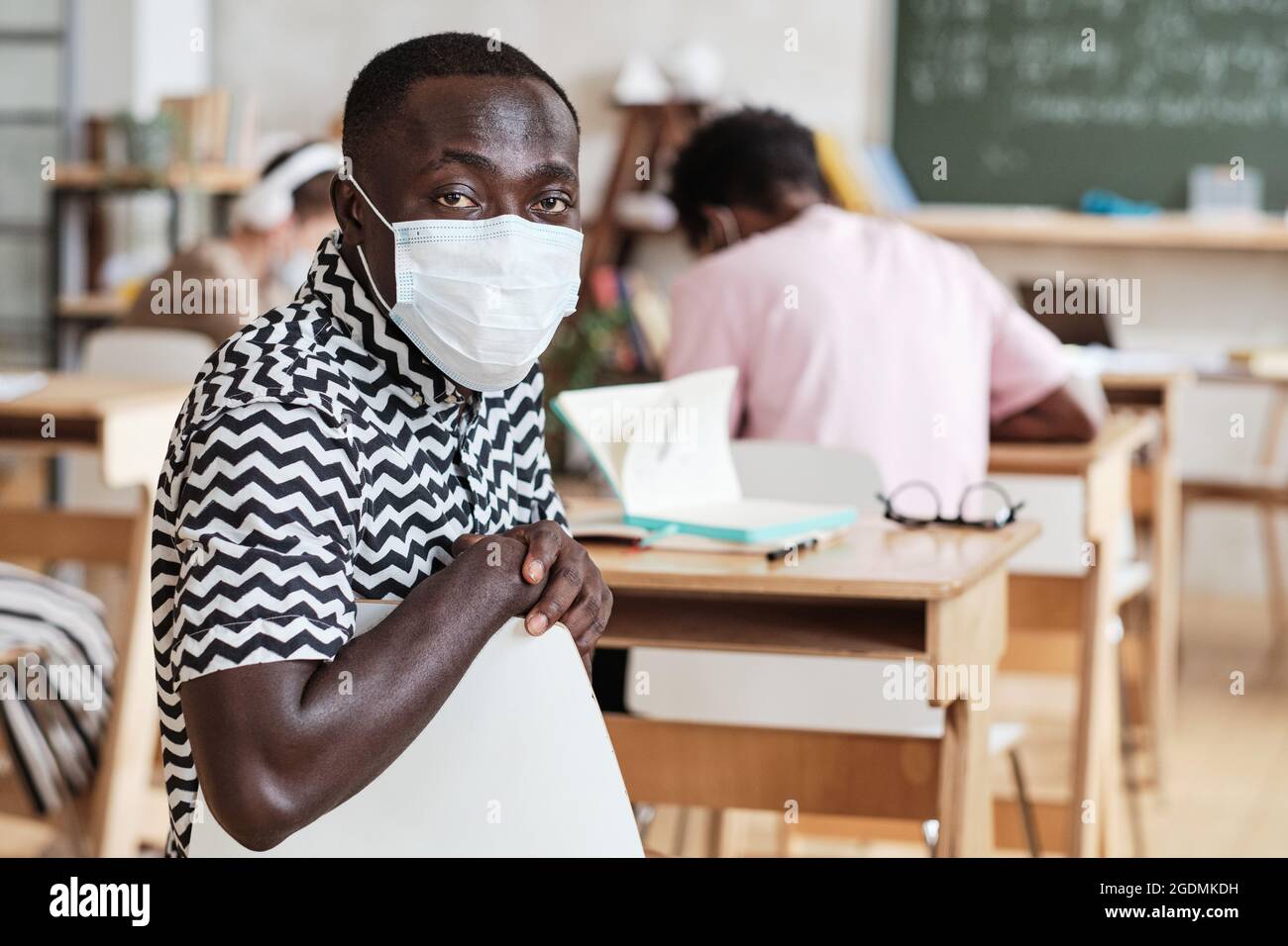 Portrait of African student in mask looking at camera while sitting at ...