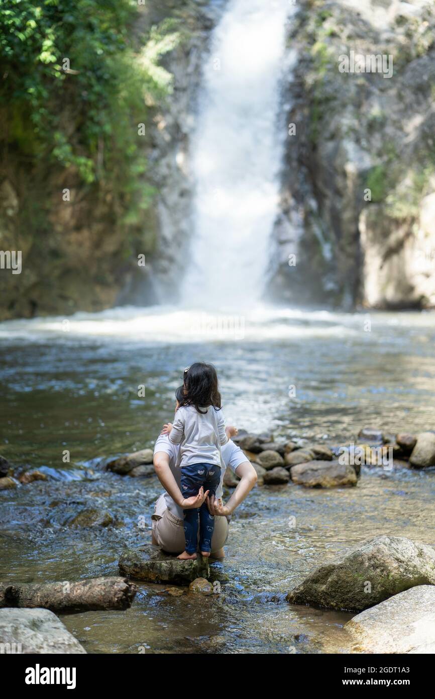 A rearview of mother and daughter sitting on the rocks with waterfalls ...