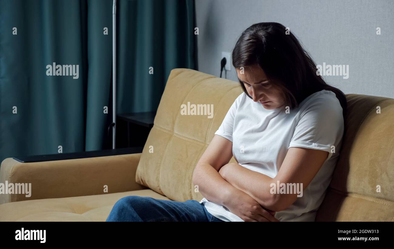 Young woman hunched over holding belly while sitting on the couch Stock ...