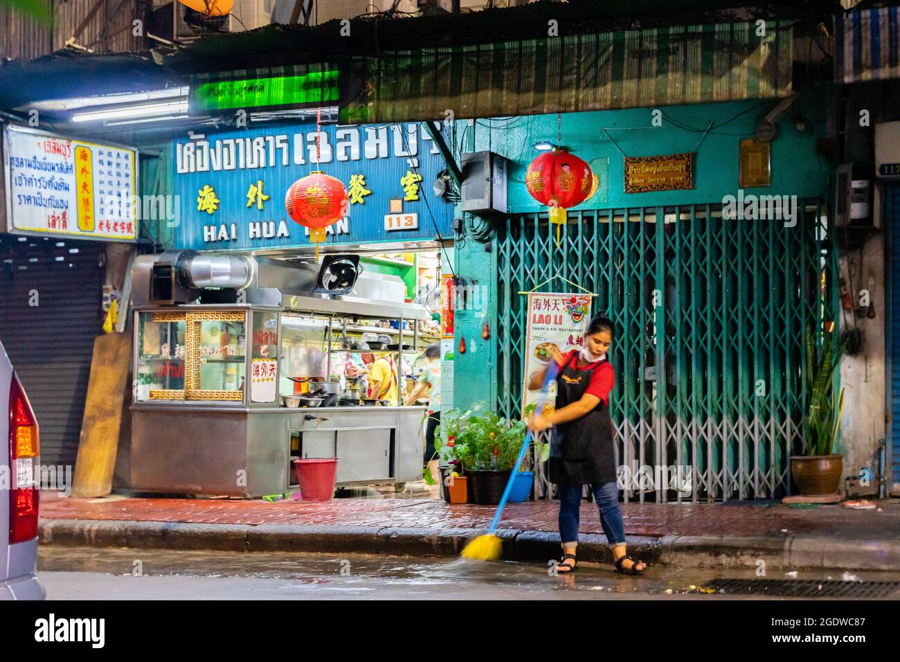 Female store staff sweeping pavement with a broom in front of the lit ...