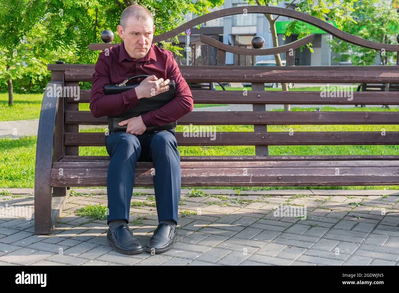 A scared man with a suitcase and a suspicious look sits on a park bench ...