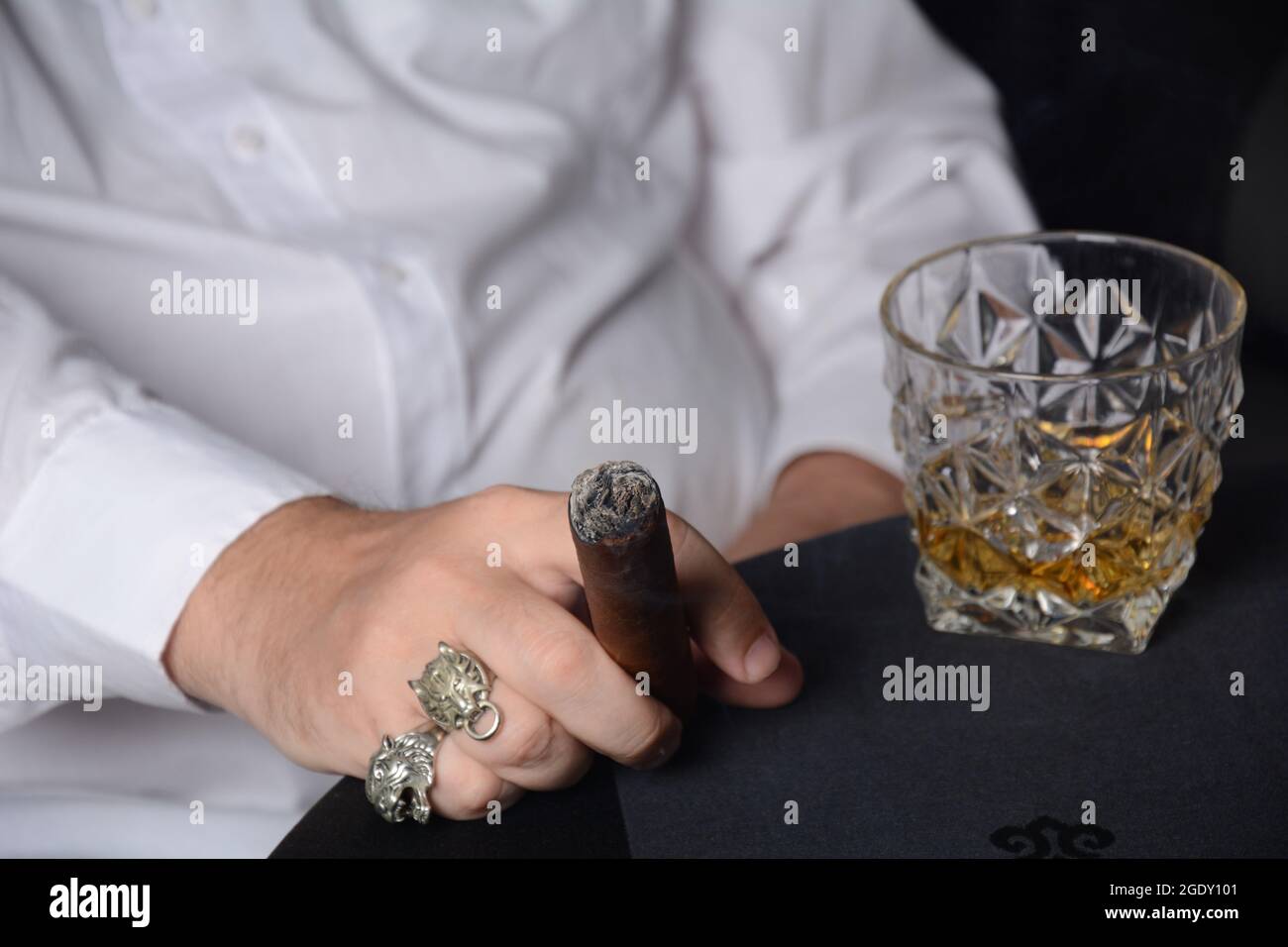 Elegant man wearing white shirt holds a smoking Cuban cigar. A glass of ...