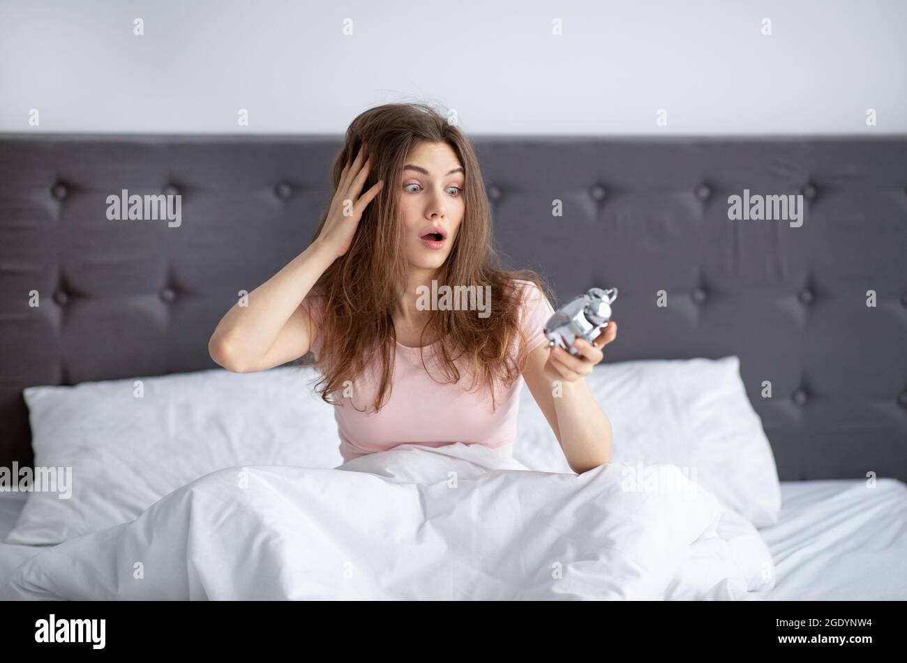 Shocked yound woman staring at alarm clock, sitting on bed at home ...