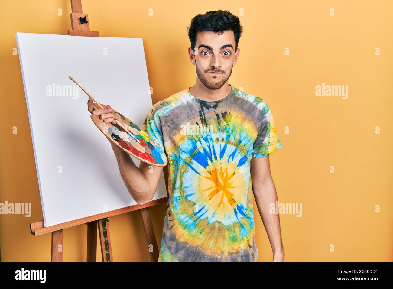 Young hispanic man standing drawing with palette by painter easel stand ...