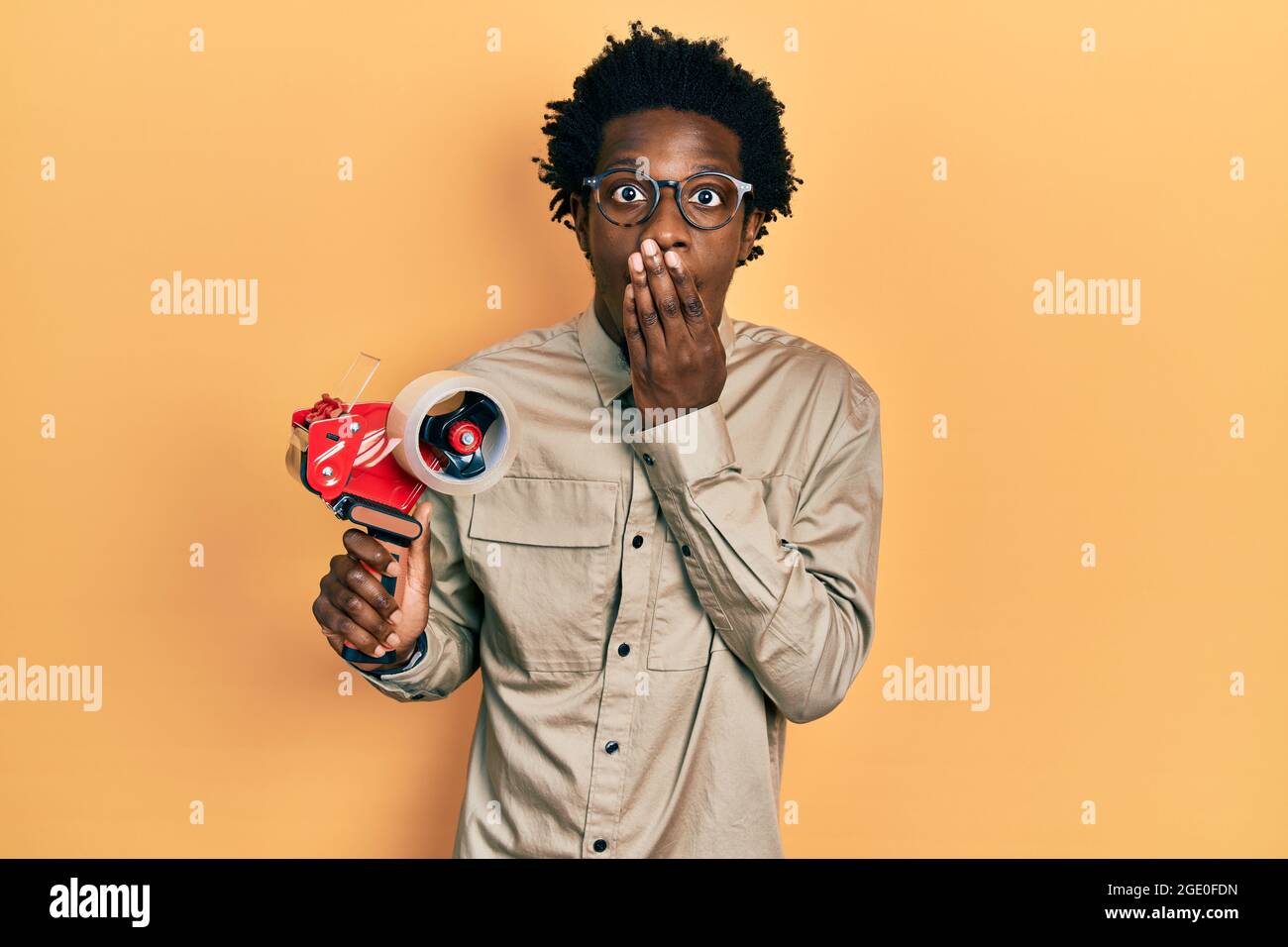 Young african american man holding packing tape machine covering mouth ...