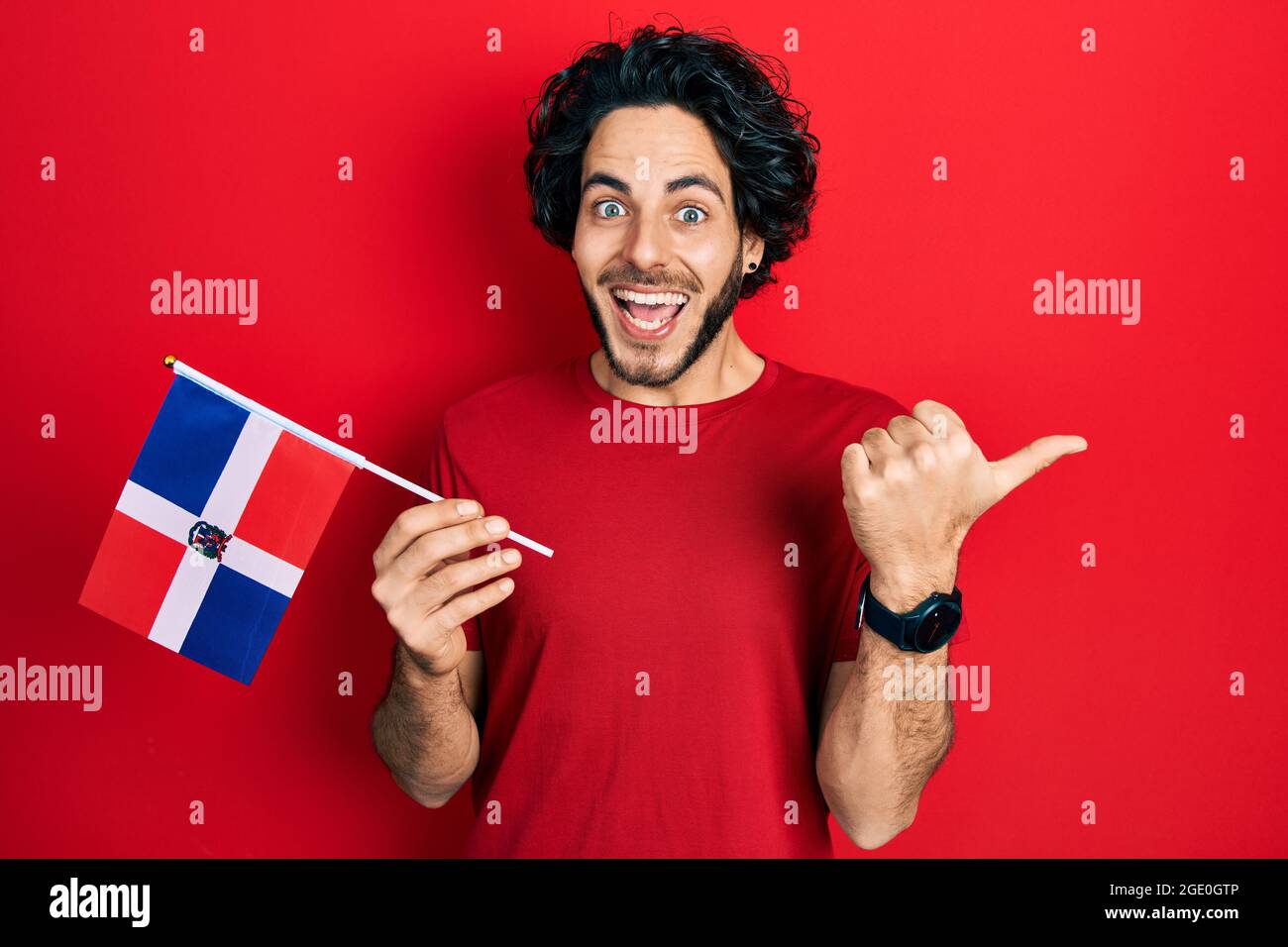 Handsome hispanic man holding dominican republic flag pointing thumb up ...
