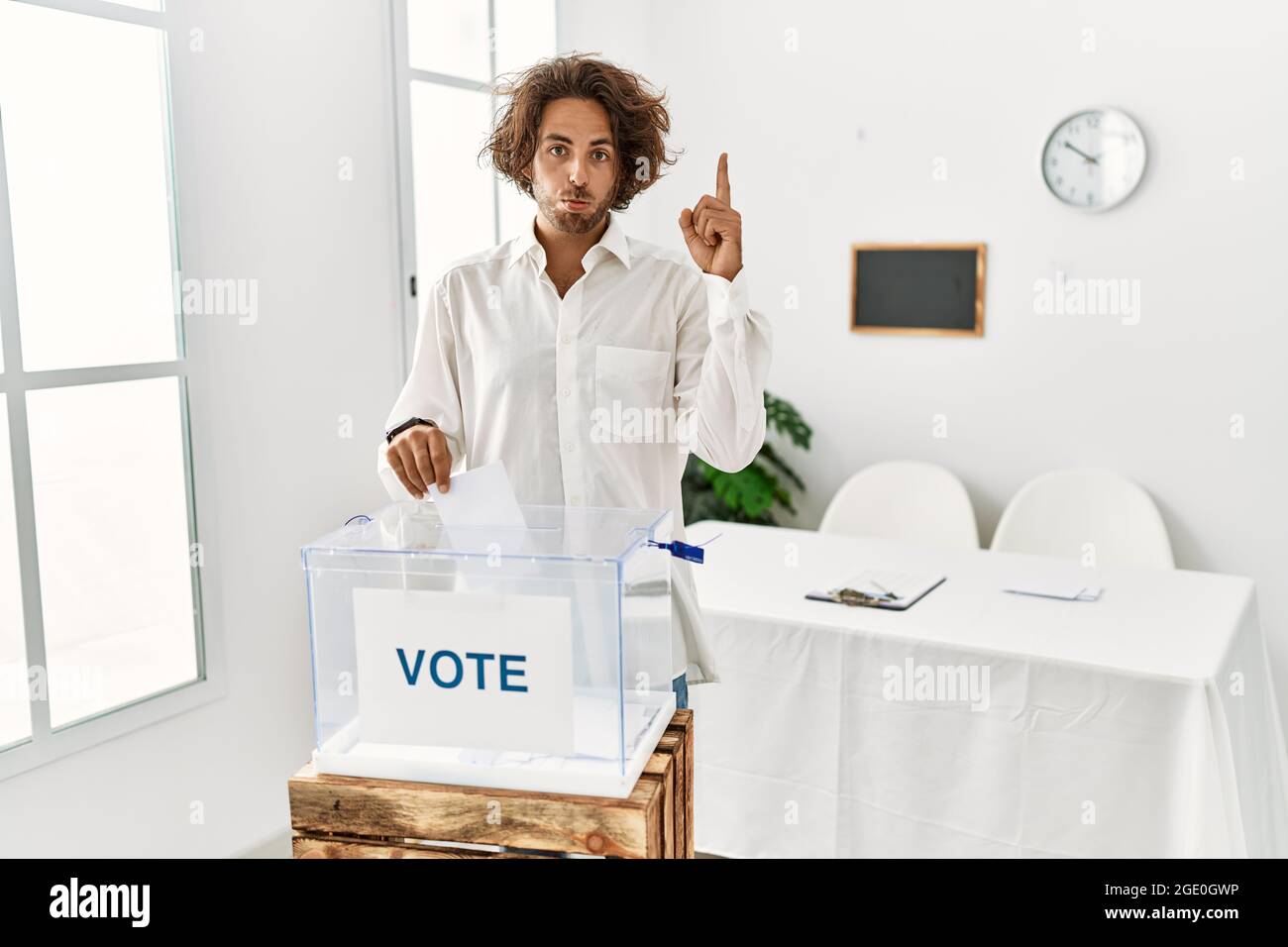 Young hispanic man voting putting envelop in ballot box pointing up ...