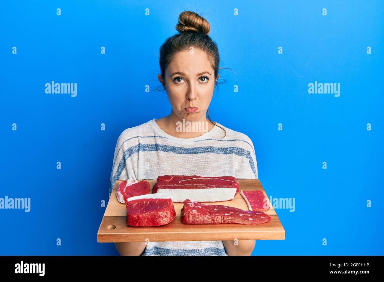 Young blonde woman holding board with raw meat depressed and worry for ...