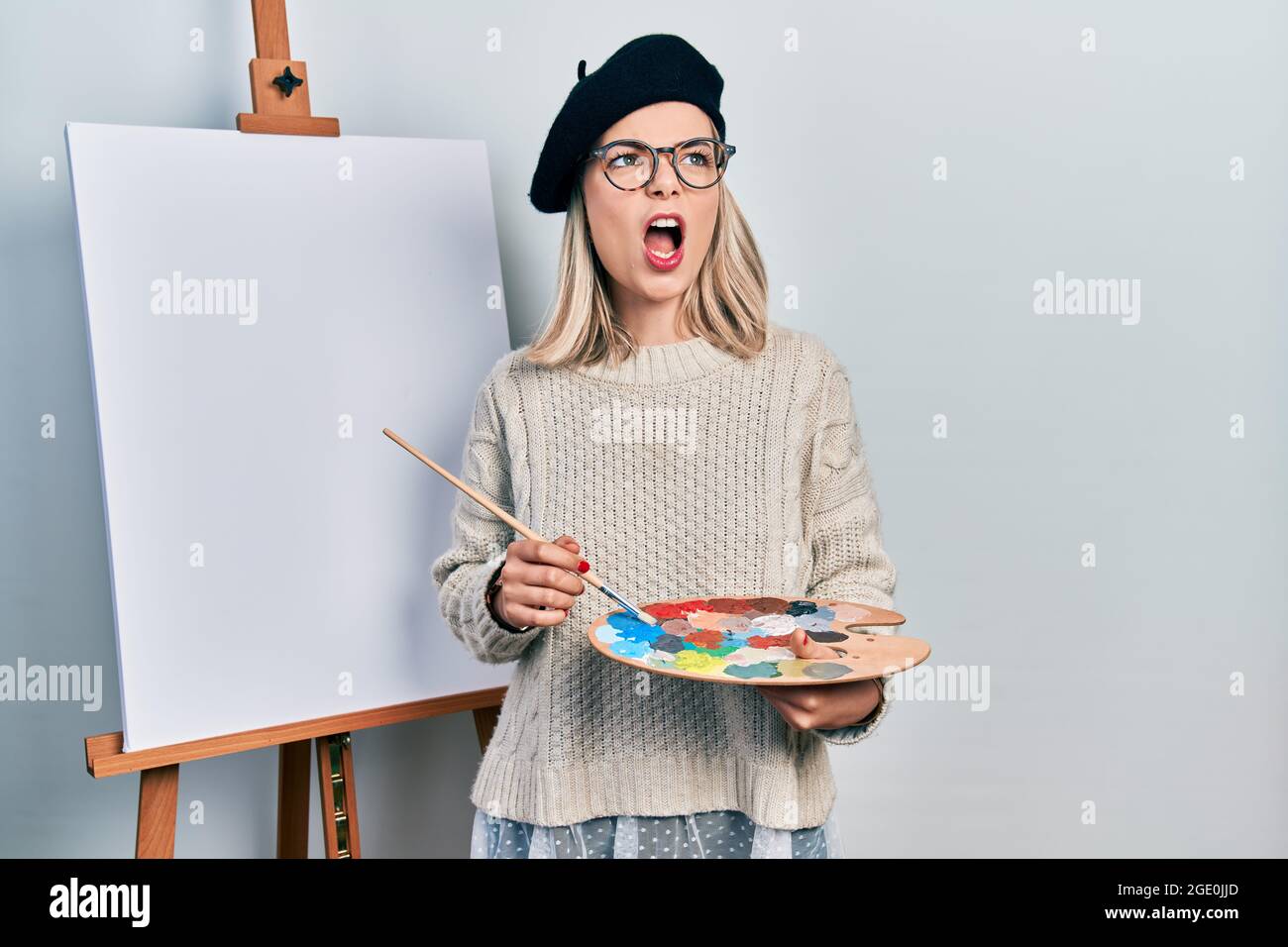 Beautiful caucasian woman drawing with palette on easel stand angry and ...