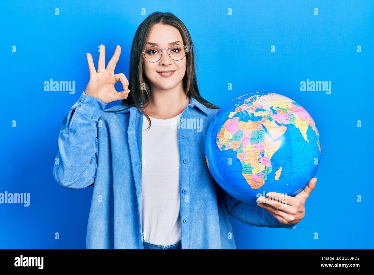 Young hispanic girl holding world ball doing ok sign with fingers ...