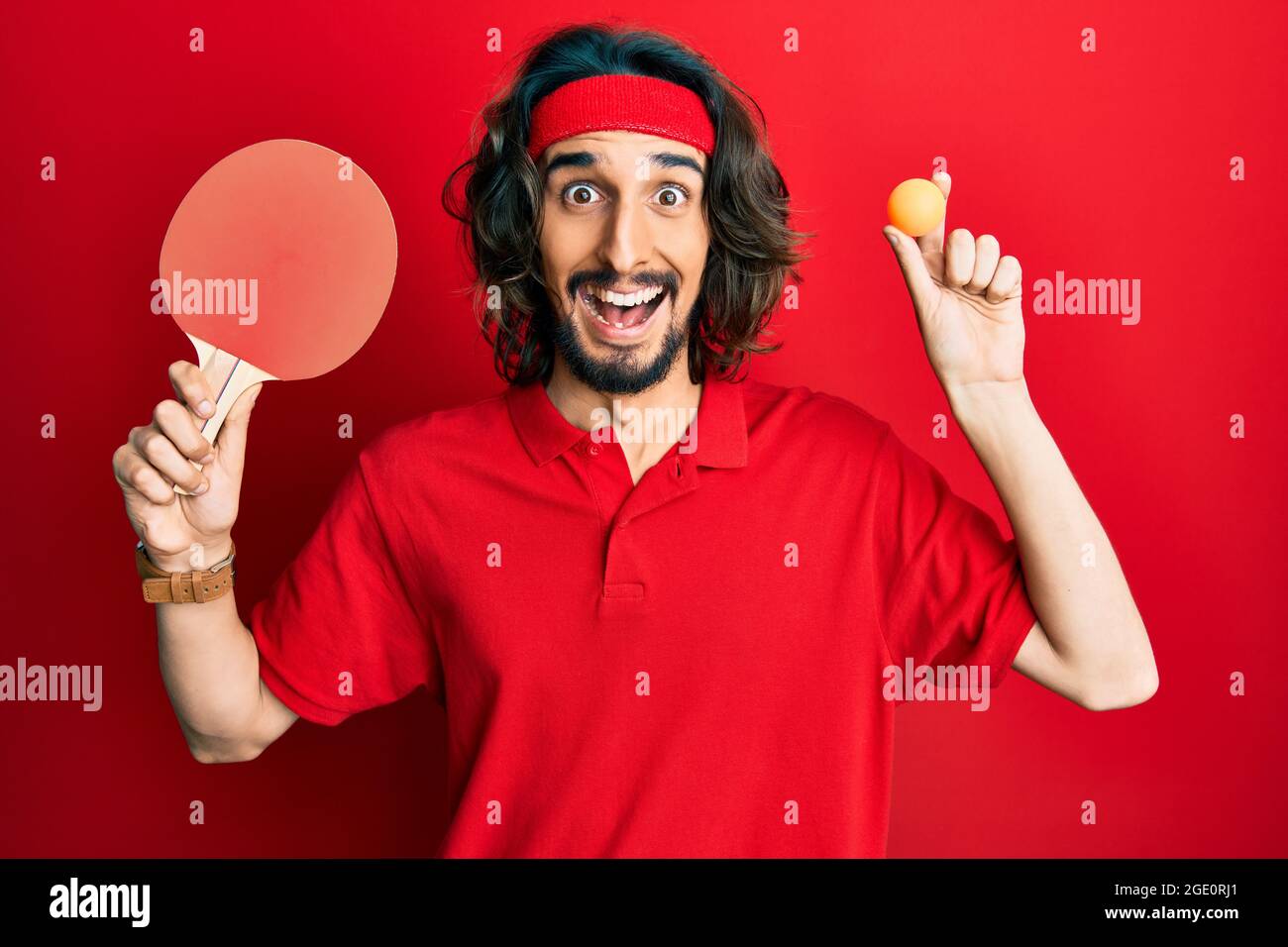 Young hispanic man holding red ping pong racket and ball celebrating ...