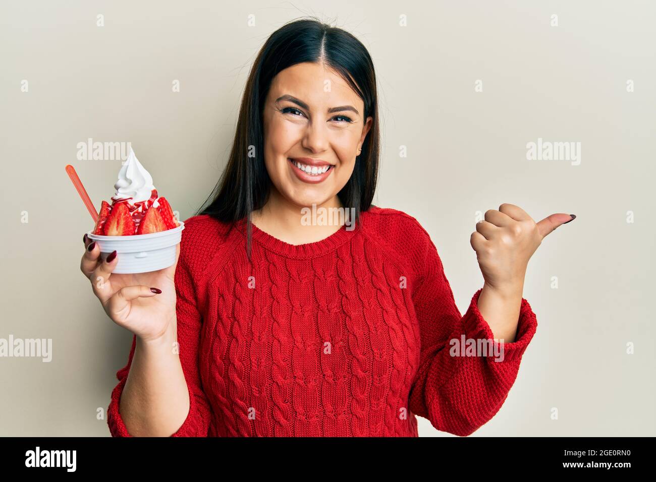 Beautiful brunette woman eating strawberry ice cream pointing thumb up ...