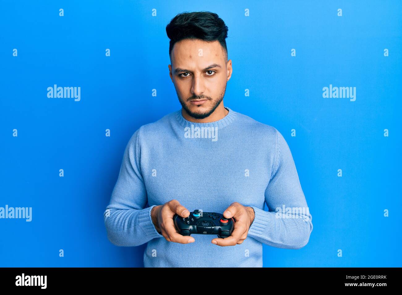 Young arab man playing video game holding controller skeptic and ...