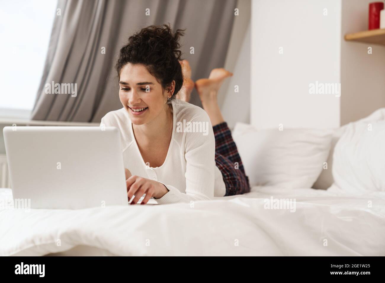 The happy typing woman lying in pajamas on the bed in a light room ...
