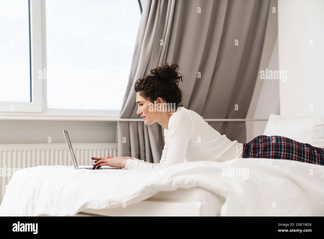 The side view of a typing girl while lying in pajamas on a bed in a ...