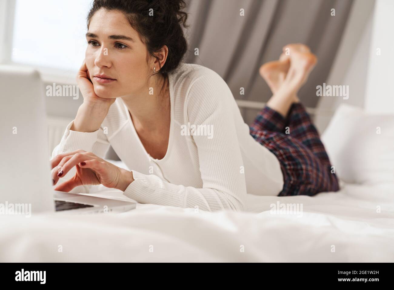 The thoughtful typing woman lying in pajamas on the bed in a light room ...