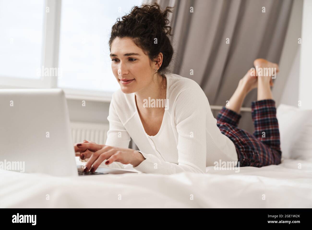 The joyful typing woman lying in pajamas on the bed in a light room ...