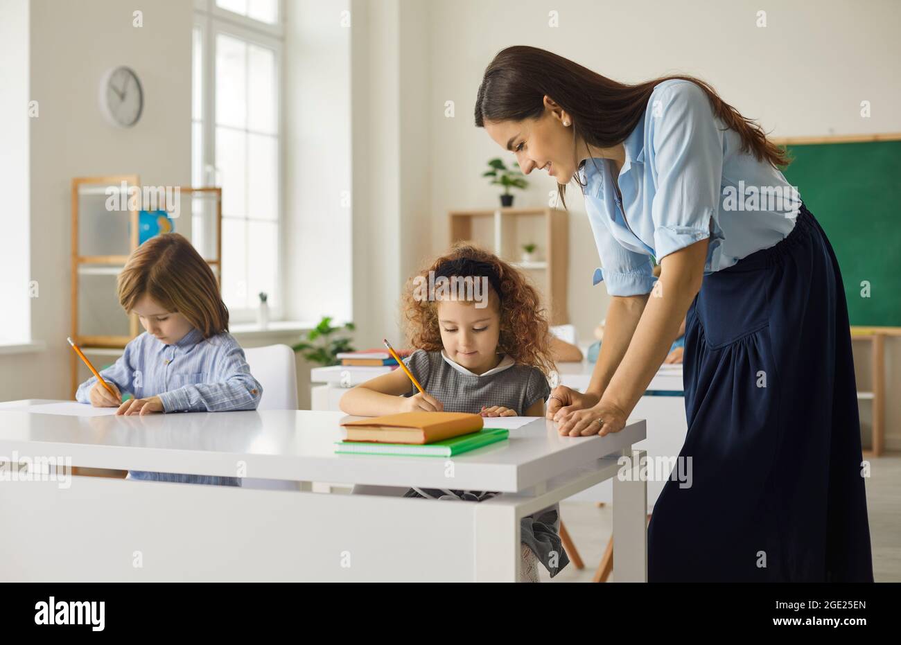 Friendly Caucasian teacher helps the junior students to complete the ...