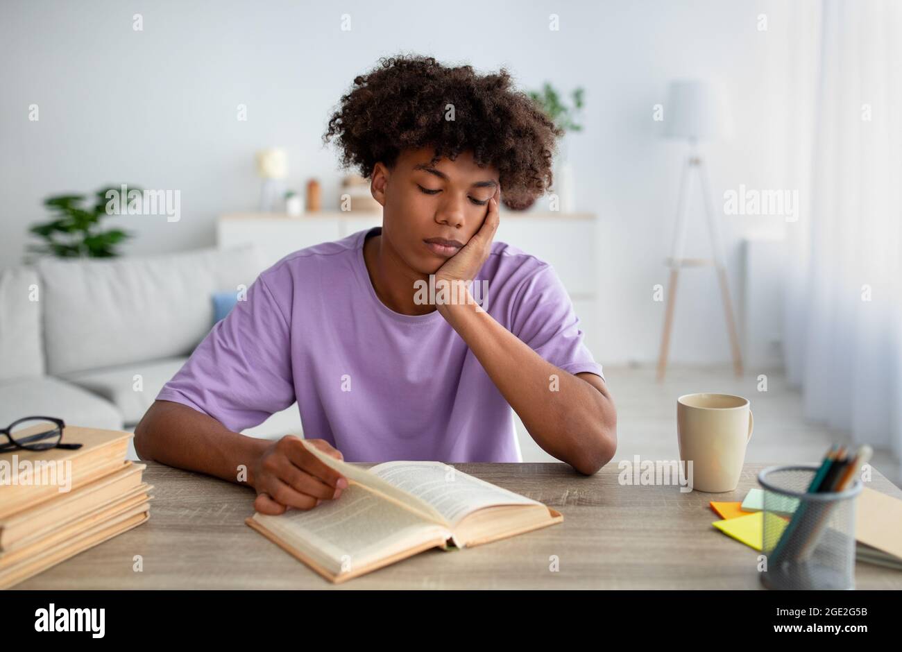 Bored black teenager sitting at desk, reading book, studying at home ...