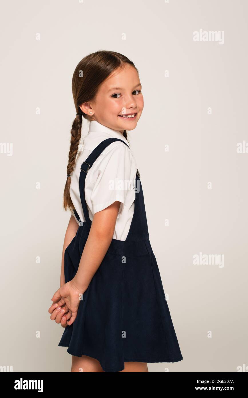 positive schoolchild standing with hands behind back and looking at ...