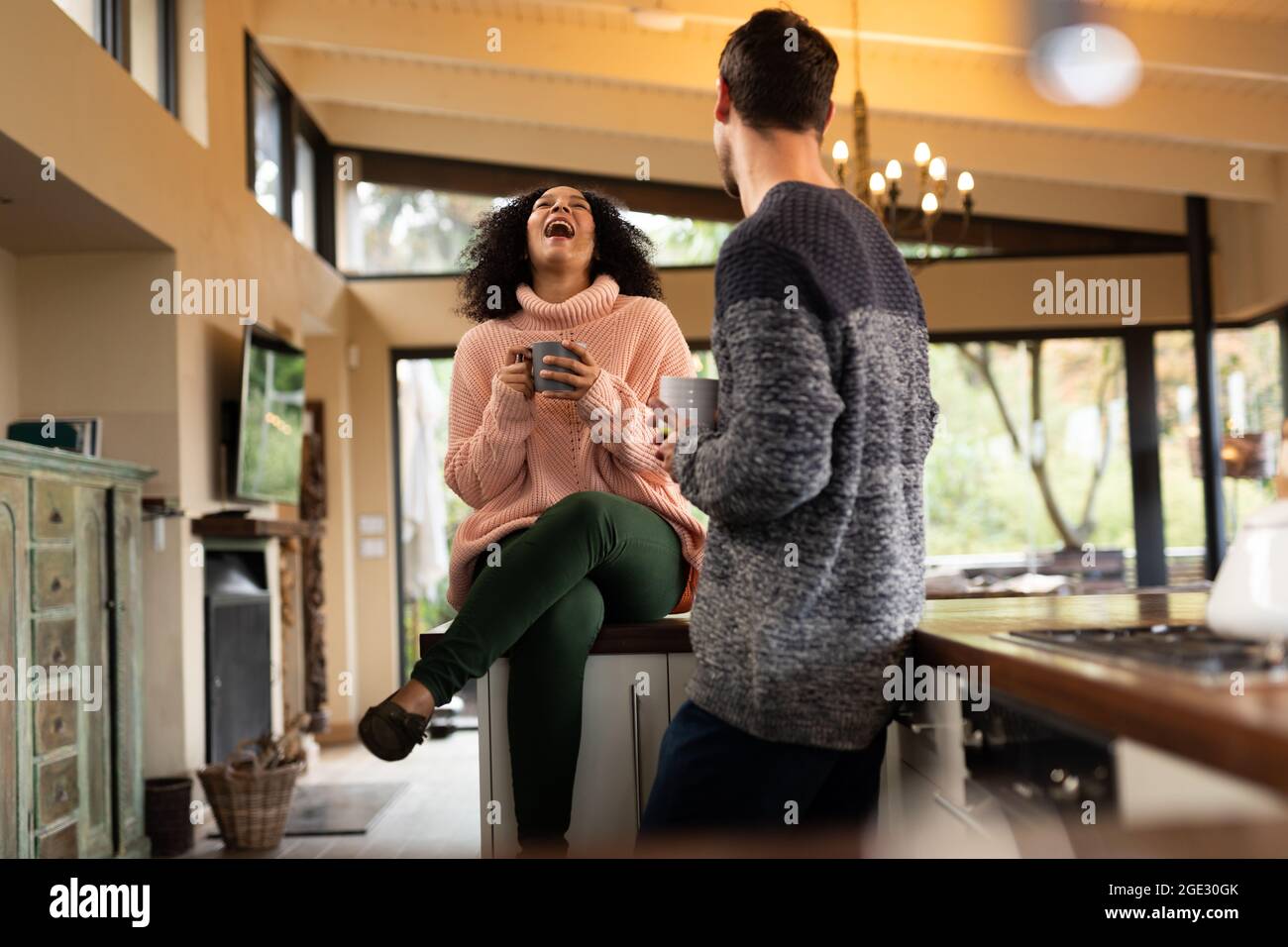 Happy diverse couple in kitchen sitting on countertop drinking coffee ...