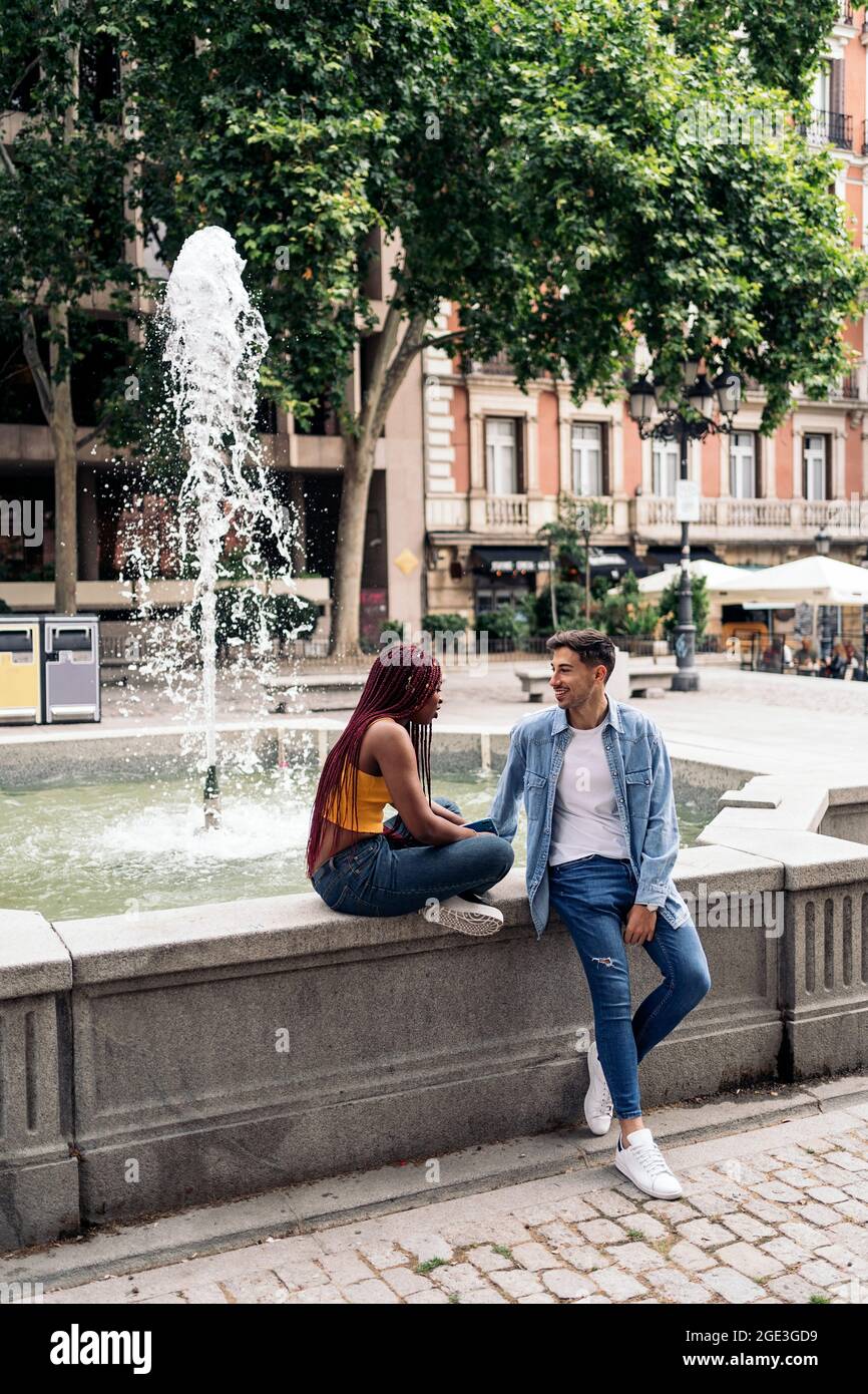 Multicultural young friends sitting next to a fountain talking and ...