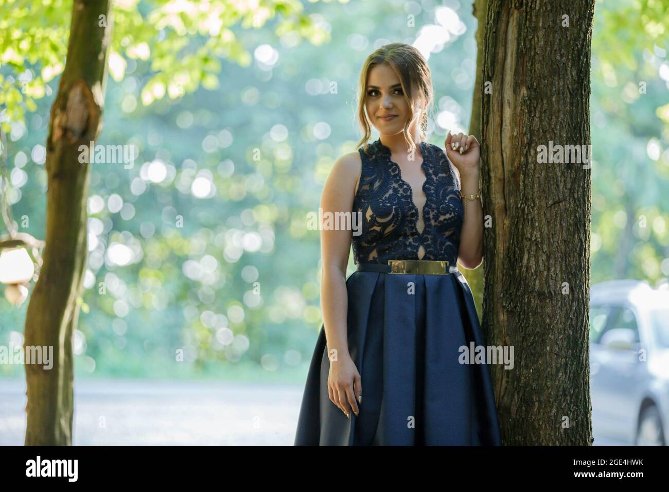 Shy Caucasian girl standing near a tree in a blue dress and looking ...