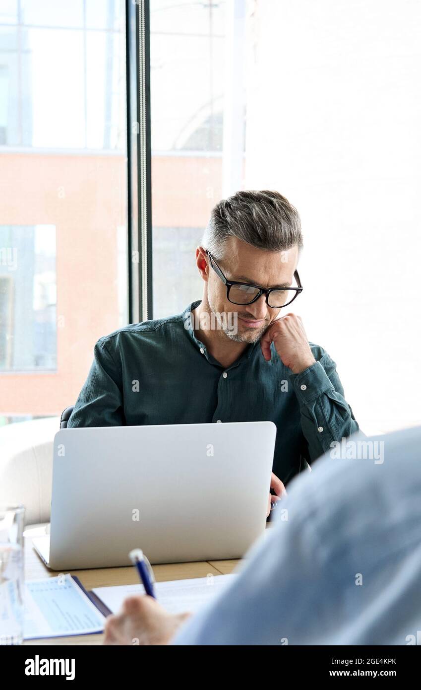 Serious male ceo looking at laptop near panoramic window at table ...
