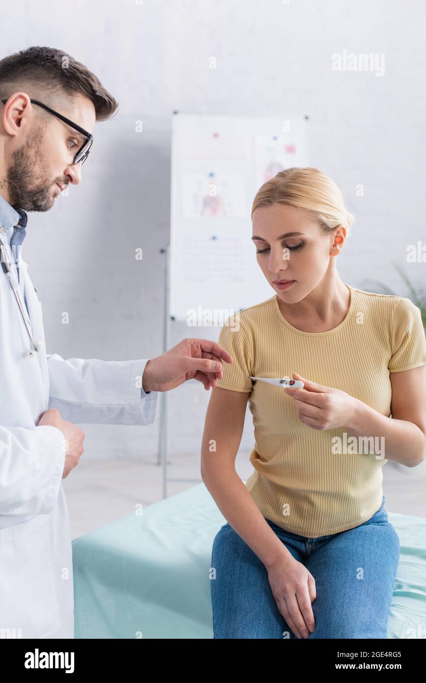 doctor giving electronic thermometer to woman sitting on medical couch ...