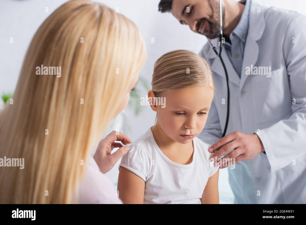 Sad kid sitting near mother and doctor with stethoscope on blurred ...