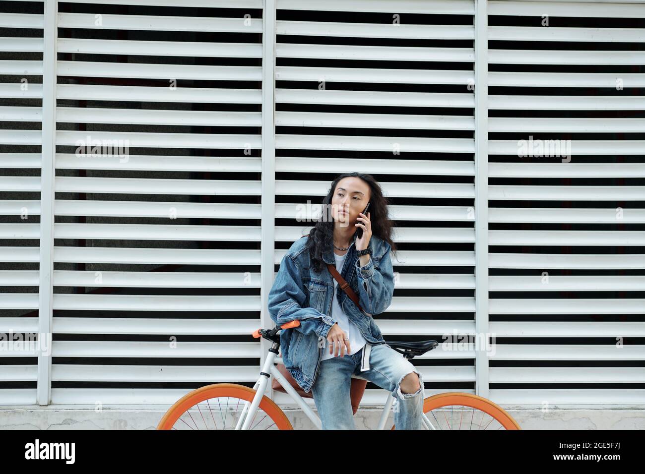 Serious young man sitting on bicycle and talking on phone with friend ...