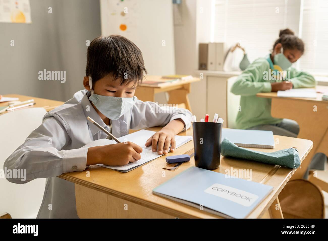 Diligent Asian schoolboy in protective mask writing in copybook against ...