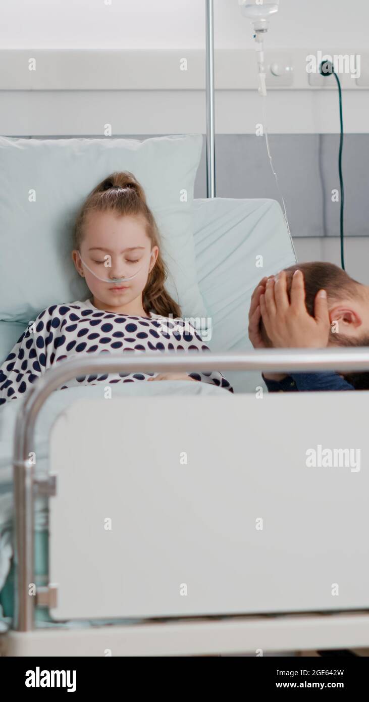 Hospitalized sick daughter sleeping after suffering medical surgery while worried father praying ...