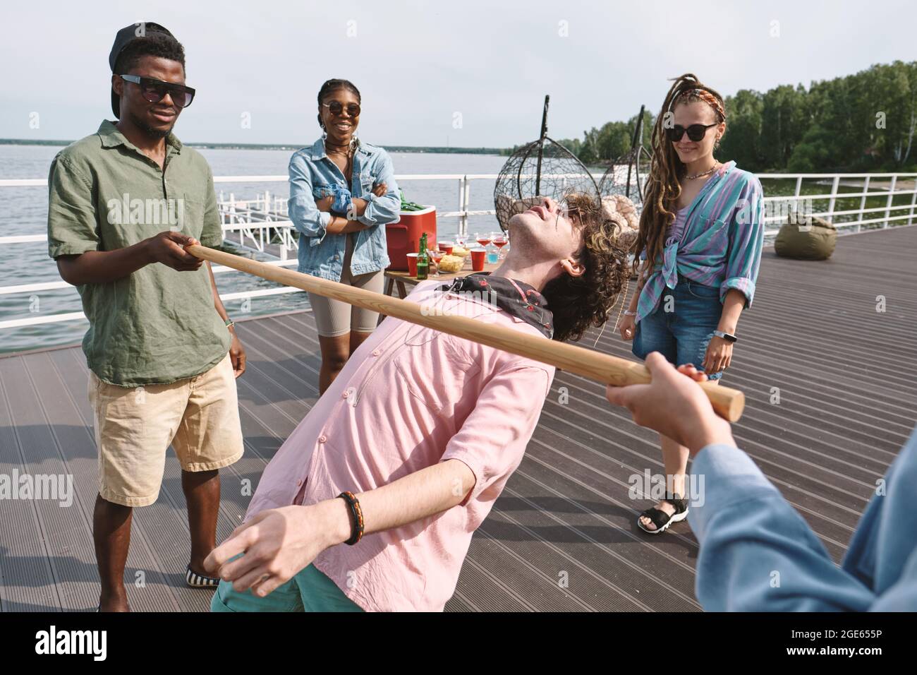 Group of joyful young people playing limbo game on pier by the lake ...