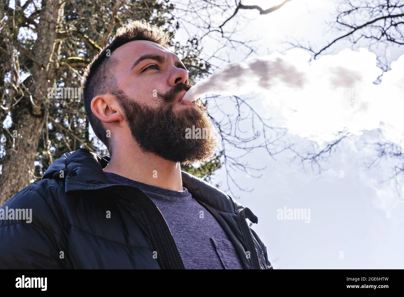 Portrait of bearded man vapes outdoors, low angle view. Large cloud of ...