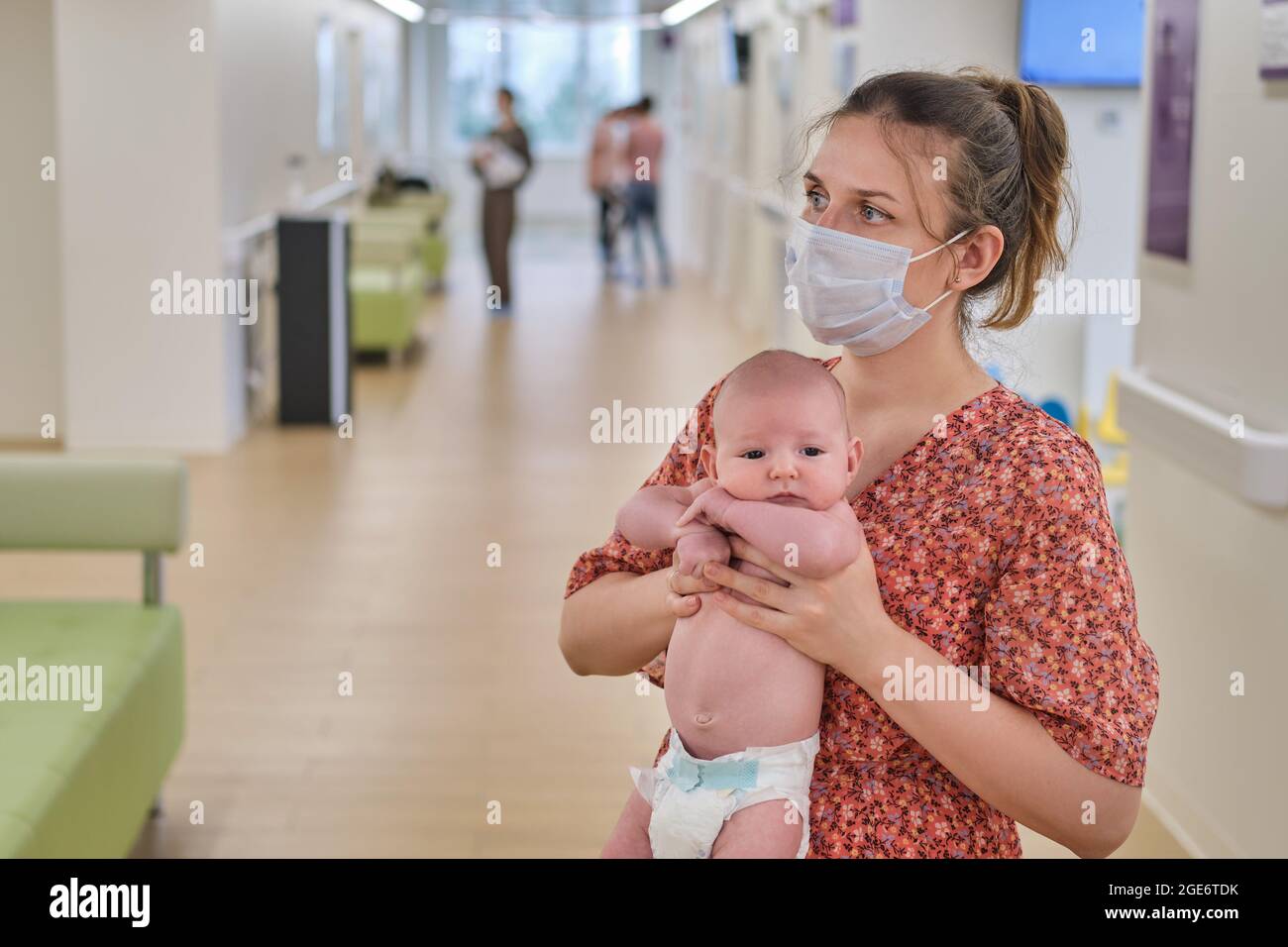 Woman mother in medical face mask holds baby boy in hospital lobby. Mom ...